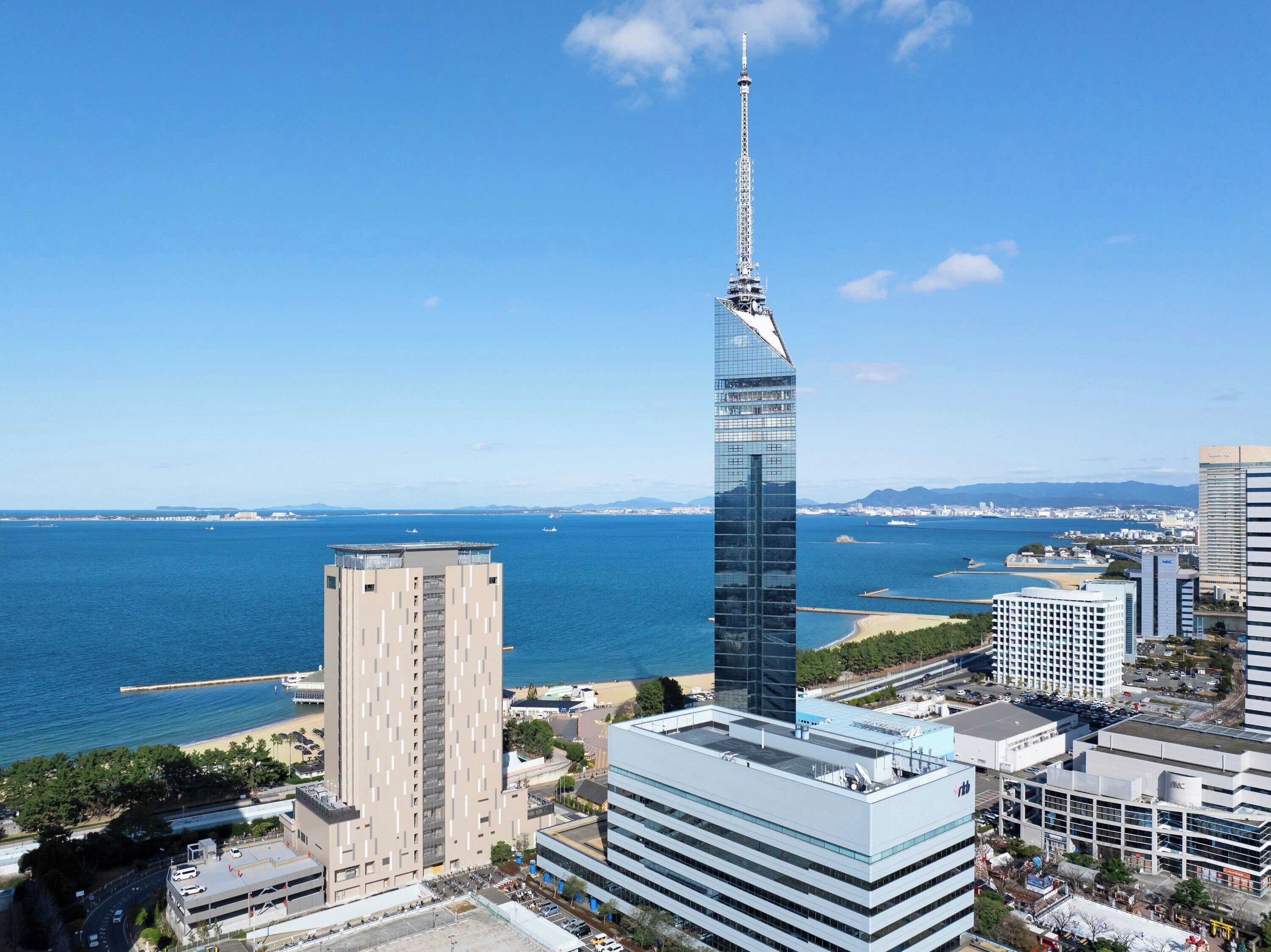 Um edifício alto com uma torre singular, cercado por outros edifícios modernos, próximo a um corpo d'água.