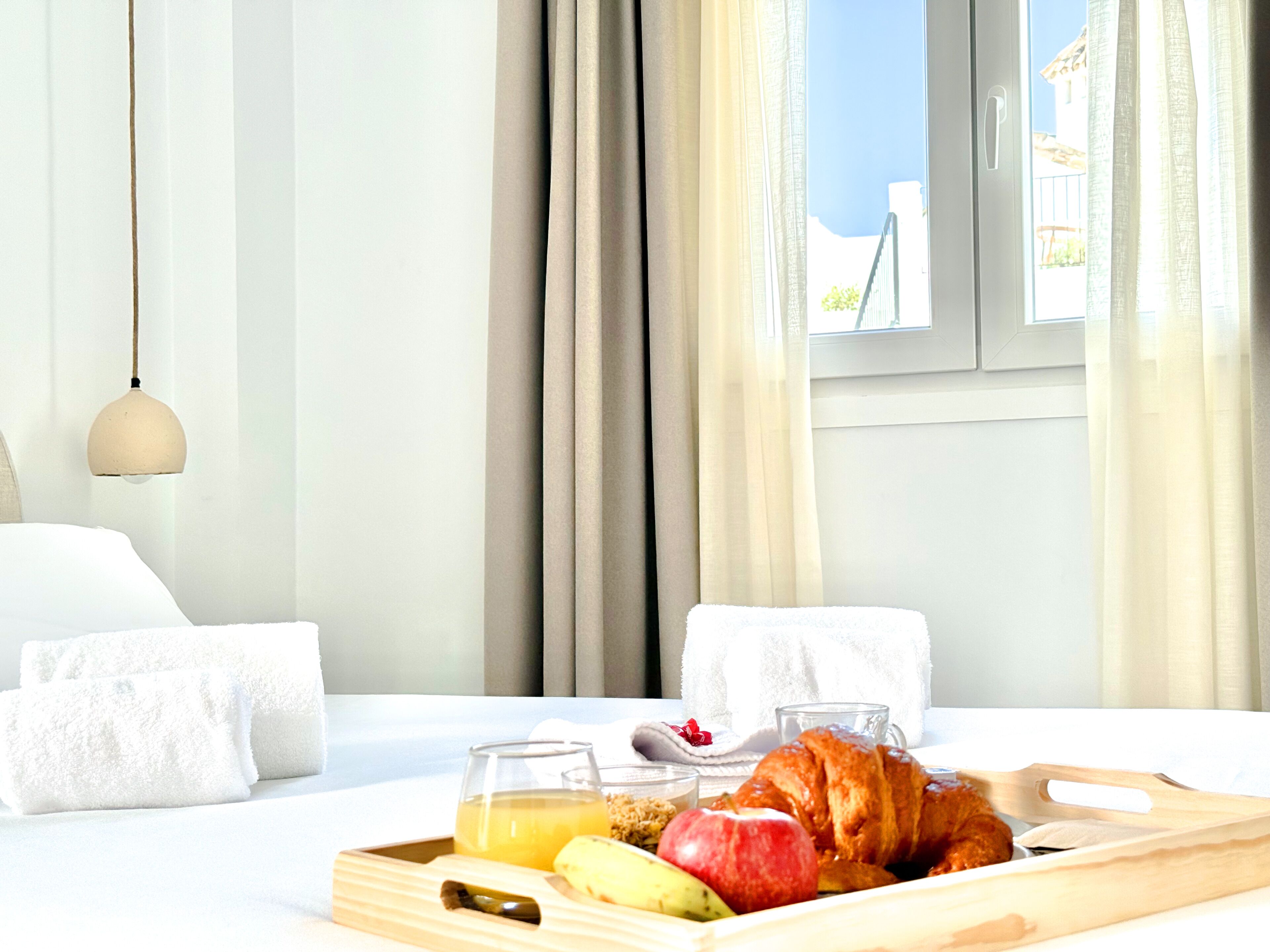 Quarto de hotel com cama, bandeja de café da manhã com croissant, maçã e suco, e vista para edifícios pela janela.