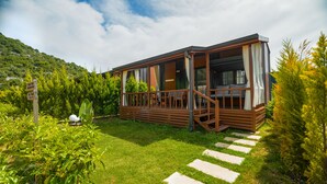 Uma cabana de madeira térrea com varanda, rodeada por um gramado bem cuidado e árvores.