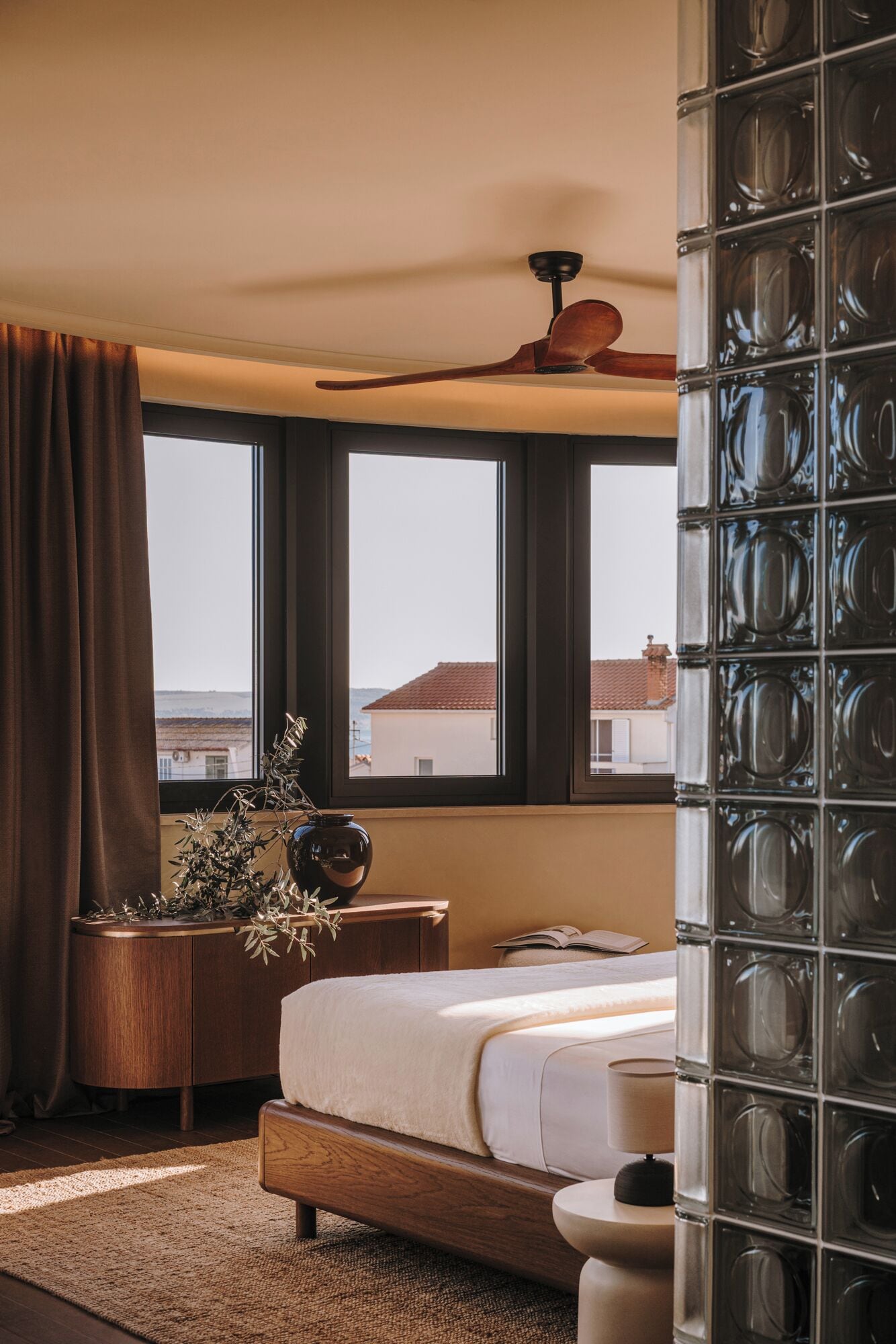 Quarto com uma grande janela, um ventilador de teto, uma mesa de cabeceira com uma planta e vista para prédios.