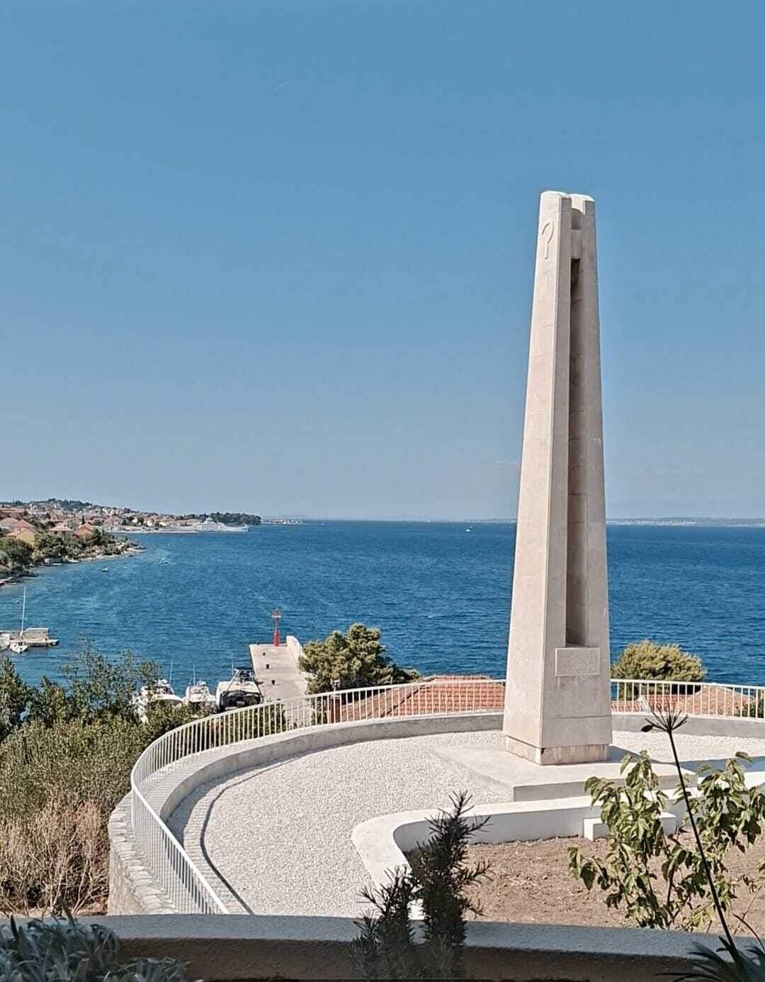 Um monumento alto com uma passarela curva em frente, com vista para uma cidade costeira e o mar.