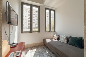Quarto com sofá, televisão na parede e duas janelas com vista para um edifício.
