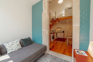 Uma sala de estar moderna com sofá cinza, paredes em tom de verde-água, piso de madeira e vista para uma área de cozinha com máquina de lavar.