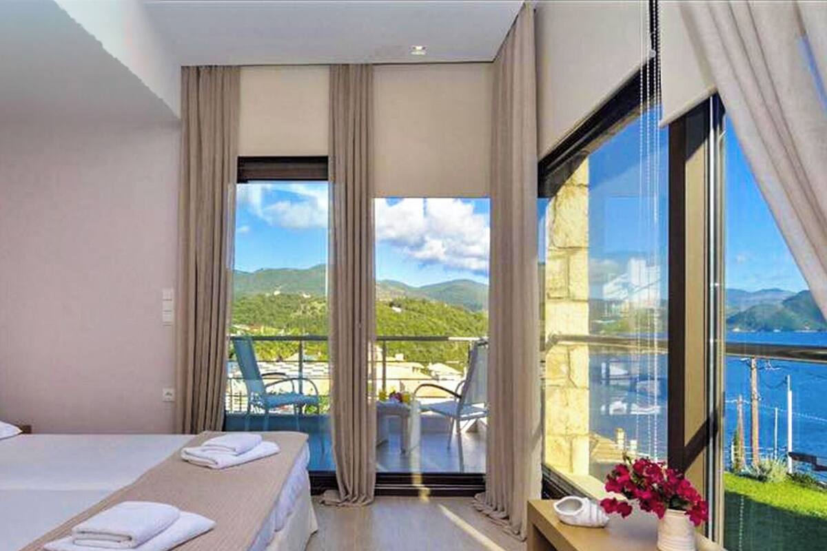 Quarto de hotel com cama, varanda com cadeiras e vista para o mar e montanhas.