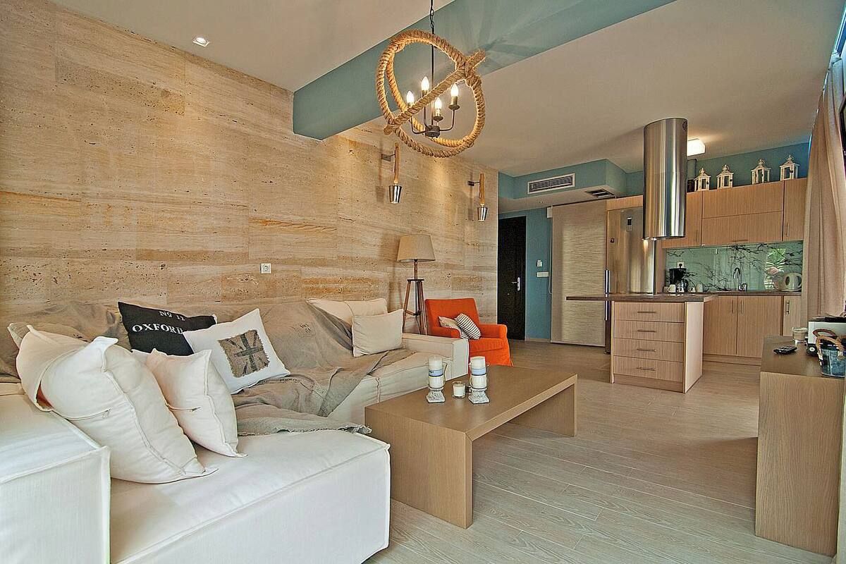 Uma sala de estar moderna com uma parede de madeira, um sofá com almofadas, uma mesa de centro e uma área de cozinha com armários e um lustre.