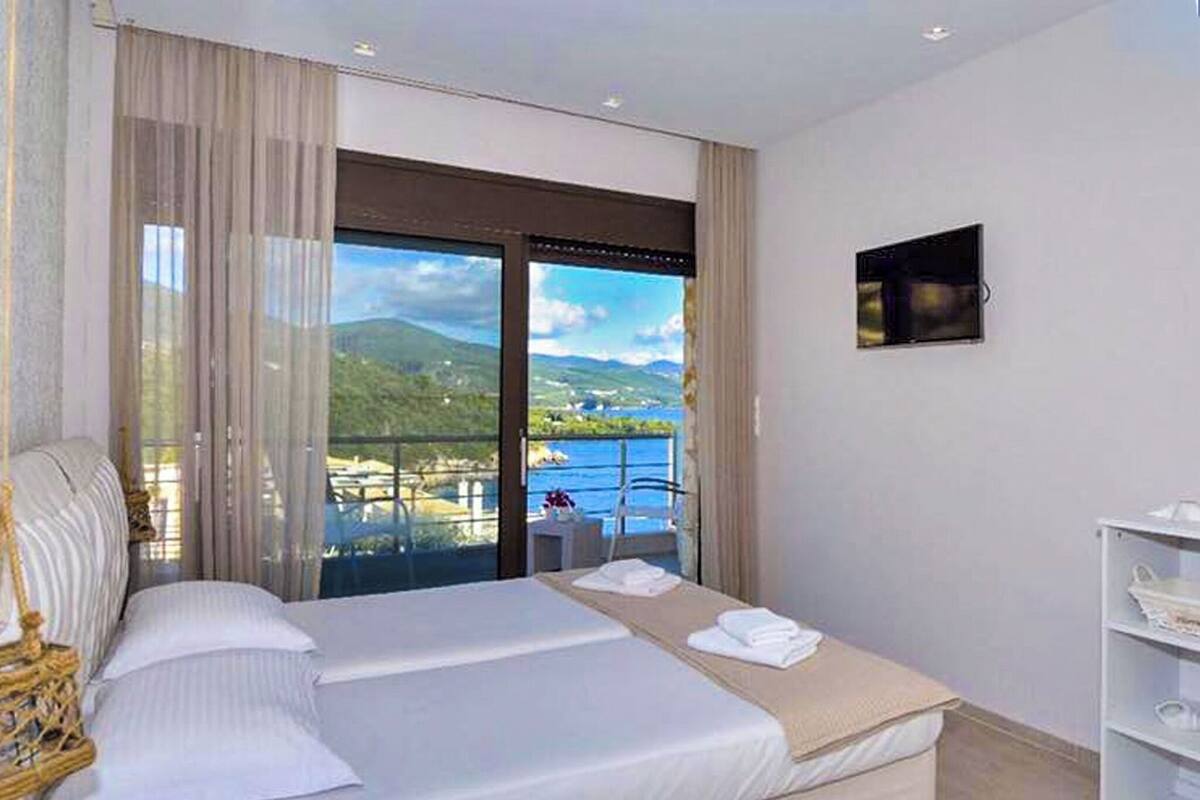 Quarto de hotel com uma cama grande, uma televisão, uma varanda com vista para o mar e colinas, e um guarda-roupa branco.
