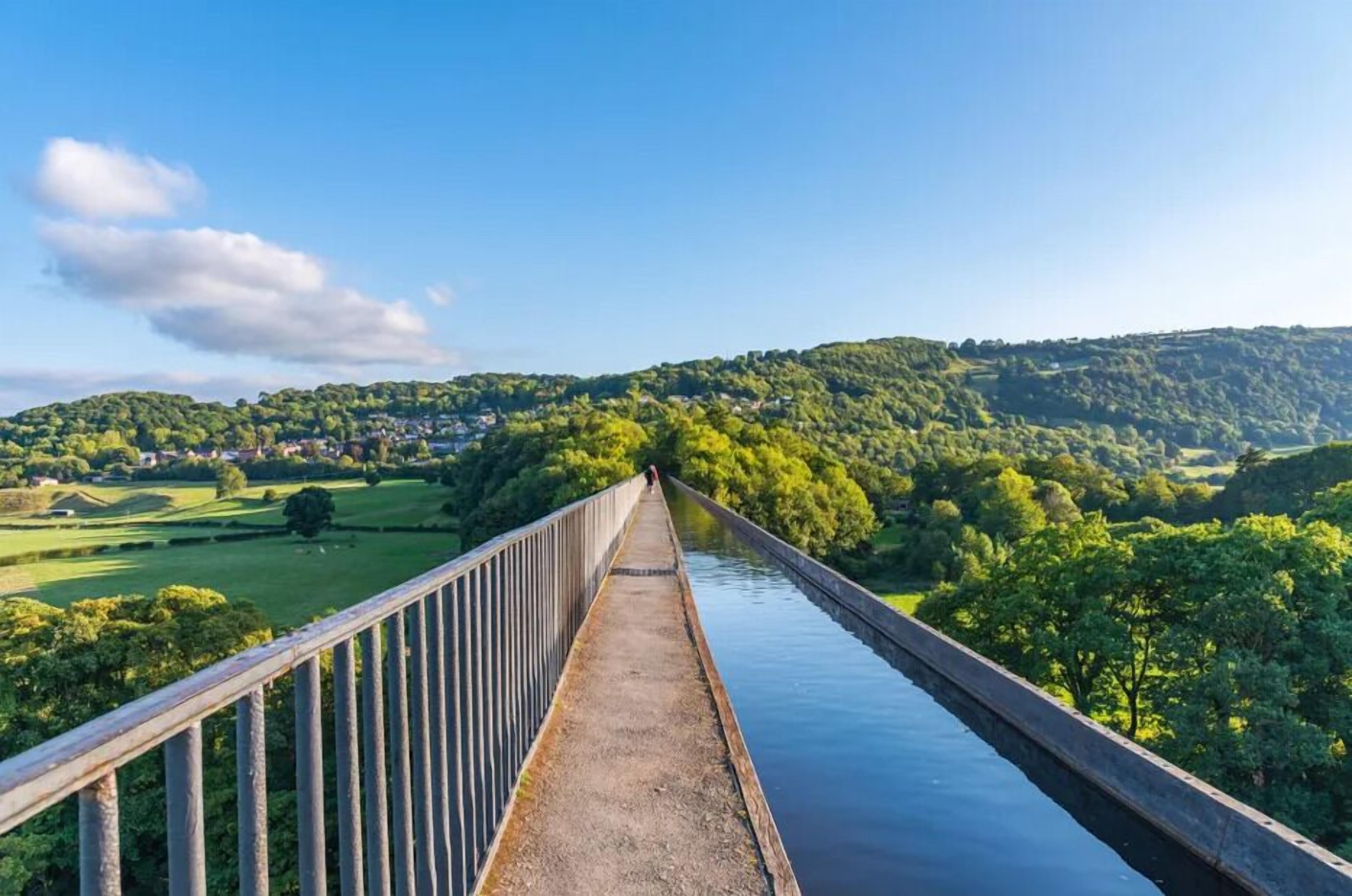Uma ponte longa e estreita, com grades de metal, que conduz a um vale verde e exuberante com edifícios dispersos.