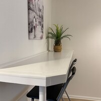 Uma mesa branca com uma cadeira preta, uma cesta de palha com uma planta e uma fotografia a preto e branco na parede.
