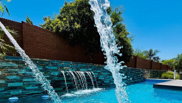 Piscina azul límpida com cascata, cercada por um muro de pedra e vegetação.