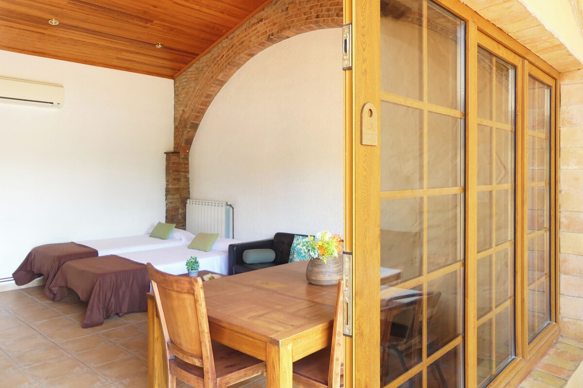 Quarto com teto de madeira, parede de tijolo, cama com almofadas, mesa com cadeiras e porta de vidro deslizante.