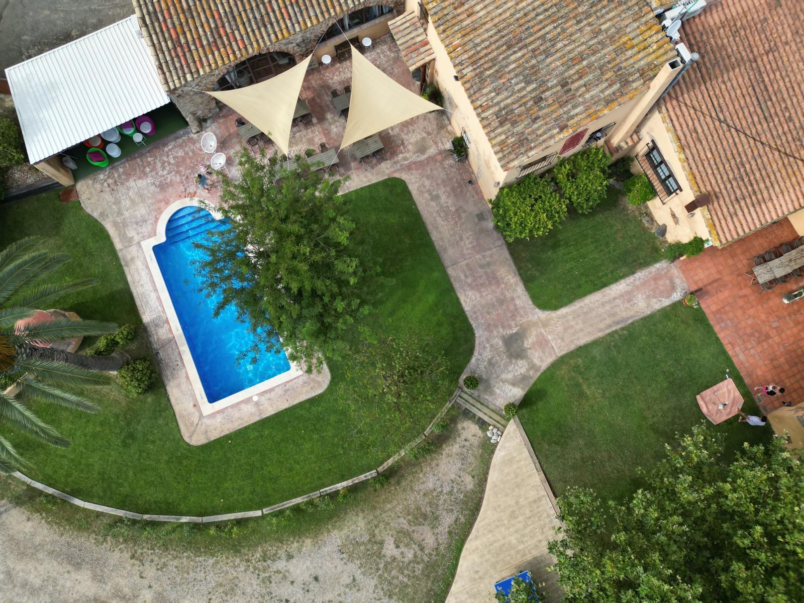 Vista aérea de uma área residencial com piscina cercada por grama, um pátio com móveis de exterior e um edifício com telhado de cerâmica.