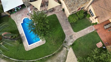 Vista aérea de uma área residencial com piscina cercada por grama, um pátio com móveis de exterior e um edifício com telhado de cerâmica.
