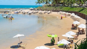 Uma praia com fileiras de guarda-sóis e espreguiçadeiras, uma faixa de areia e mar calmo.