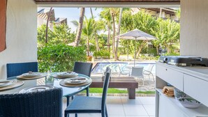 Uma área de refeições ao ar livre com vista para uma piscina, rodeada por vegetação exuberante e palmeiras.