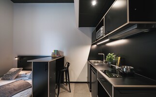 Cozinha compacta com bancada, pia e fogão. Há um pequeno balcão com banquetas e uma estante.