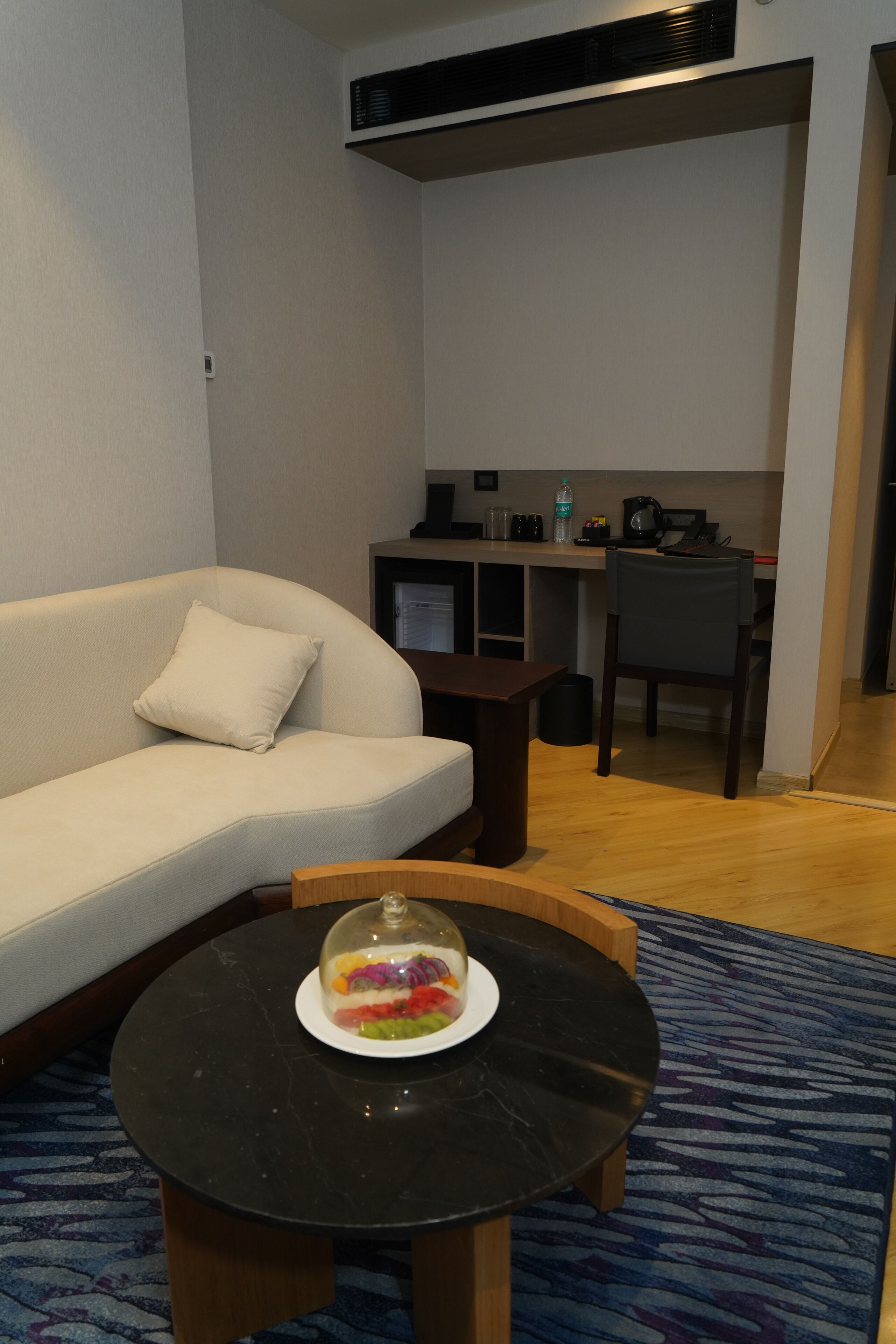Uma sala de estar com um sofá, uma mesa de centro com uma cúpula de vidro, um tapete estampado e uma cozinha compacta ao fundo.