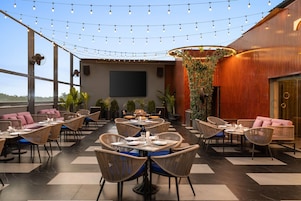 Um restaurante com mesas redondas e cadeiras de vime, luzes pendentes e uma tela grande na parede.