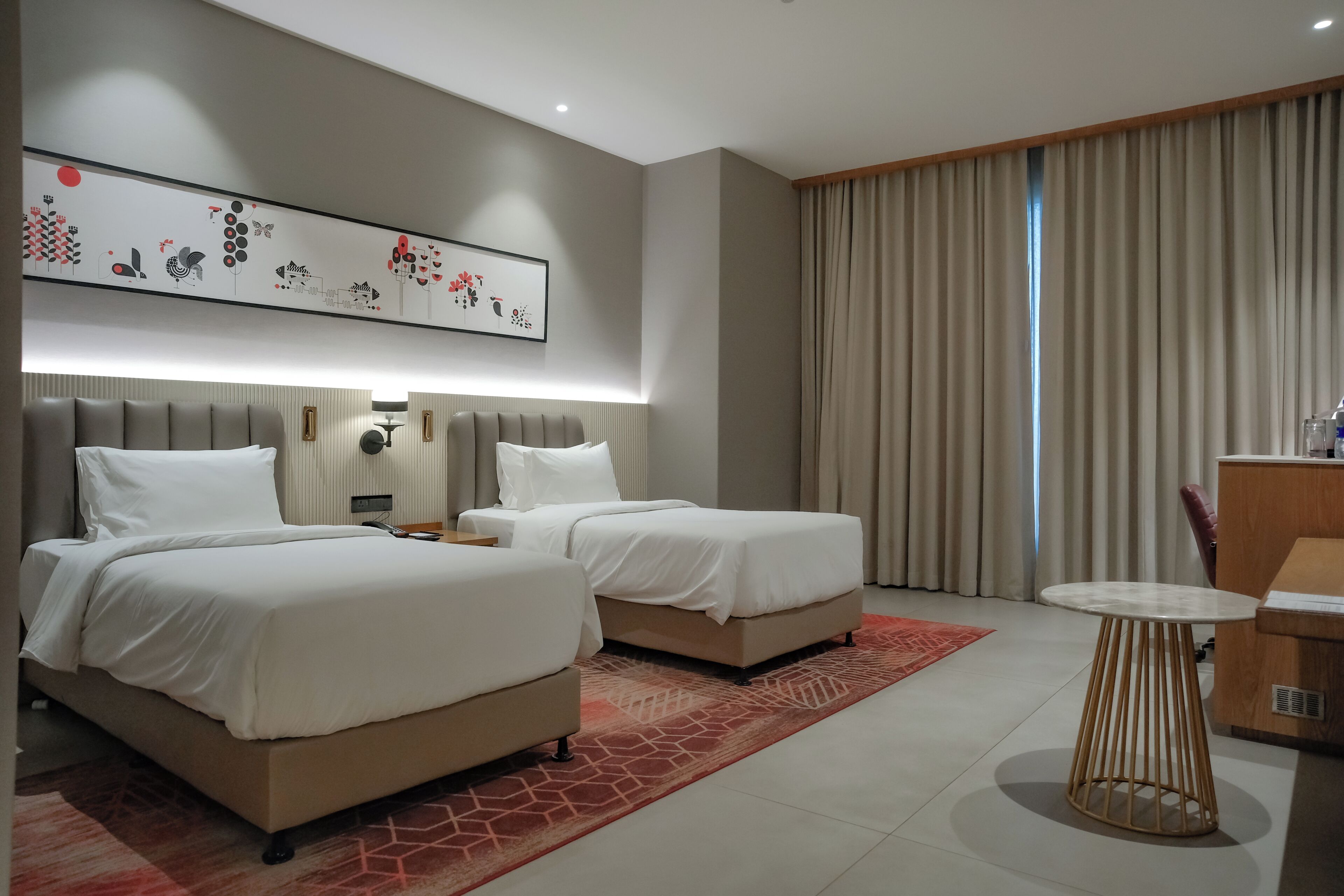 Quarto de hotel com duas camas, uma escrivaninha, uma cadeira, uma mesa redonda, um tapete com padrão vermelho e uma obra de arte na parede.