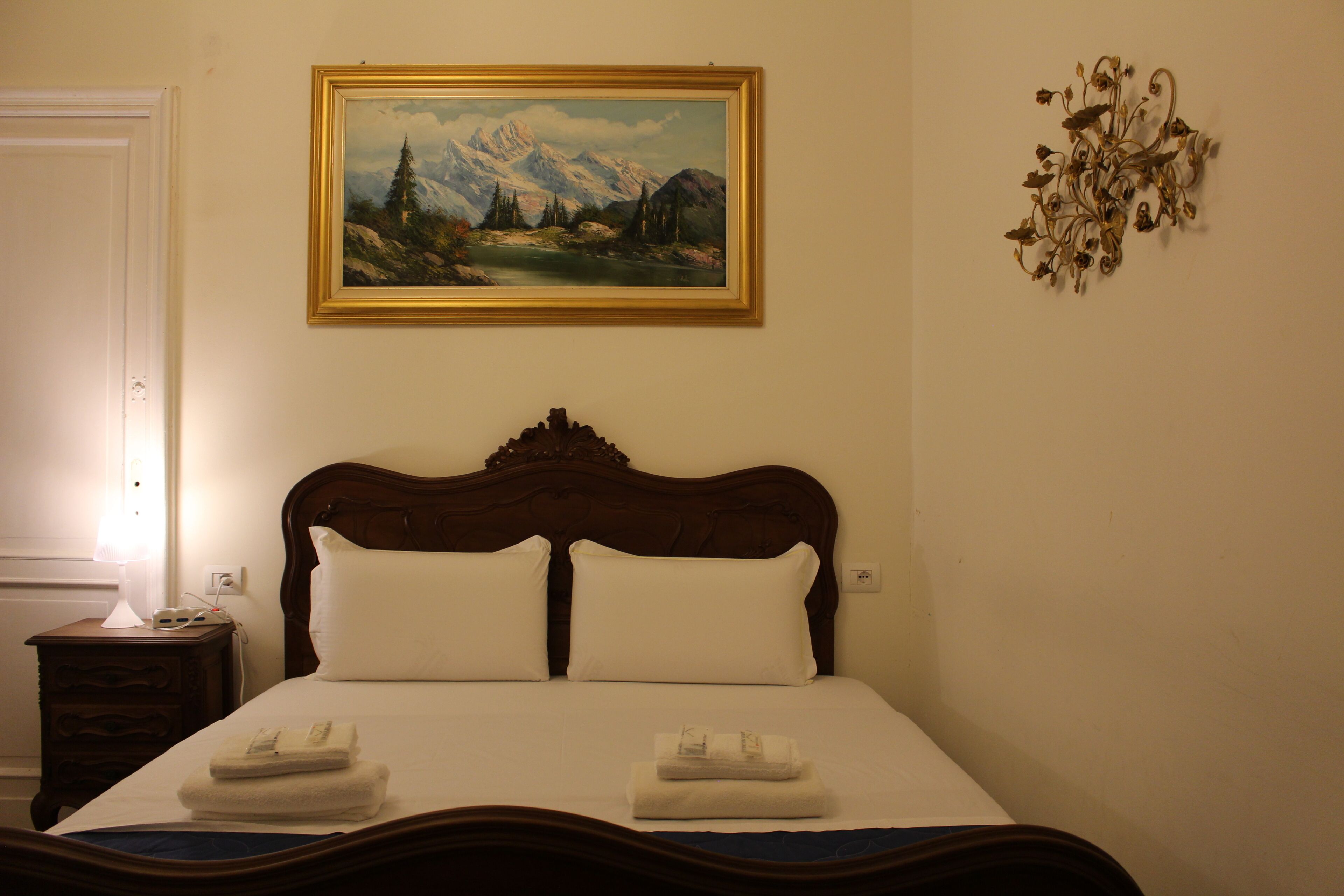 Quarto de hotel com uma cama, um quadro de montanhas, uma mesinha de cabeceira e um adorno na parede.
