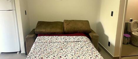 Quarto pequeno e compacto com cama, sofá, geladeira e vaso sanitário.