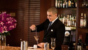 Um bartender servindo uma bebida em uma taça de martini em um bar, com uma variedade de garrafas ao fundo.