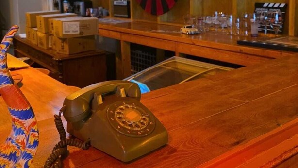 Uma área de bar com um alvo de dardos, um telefone antigo e uma tela digital exibindo informações.