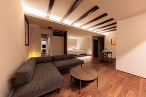 Uma sala de estar moderna com sofá seccional, mesa de centro de madeira redonda e uma cama ao fundo.