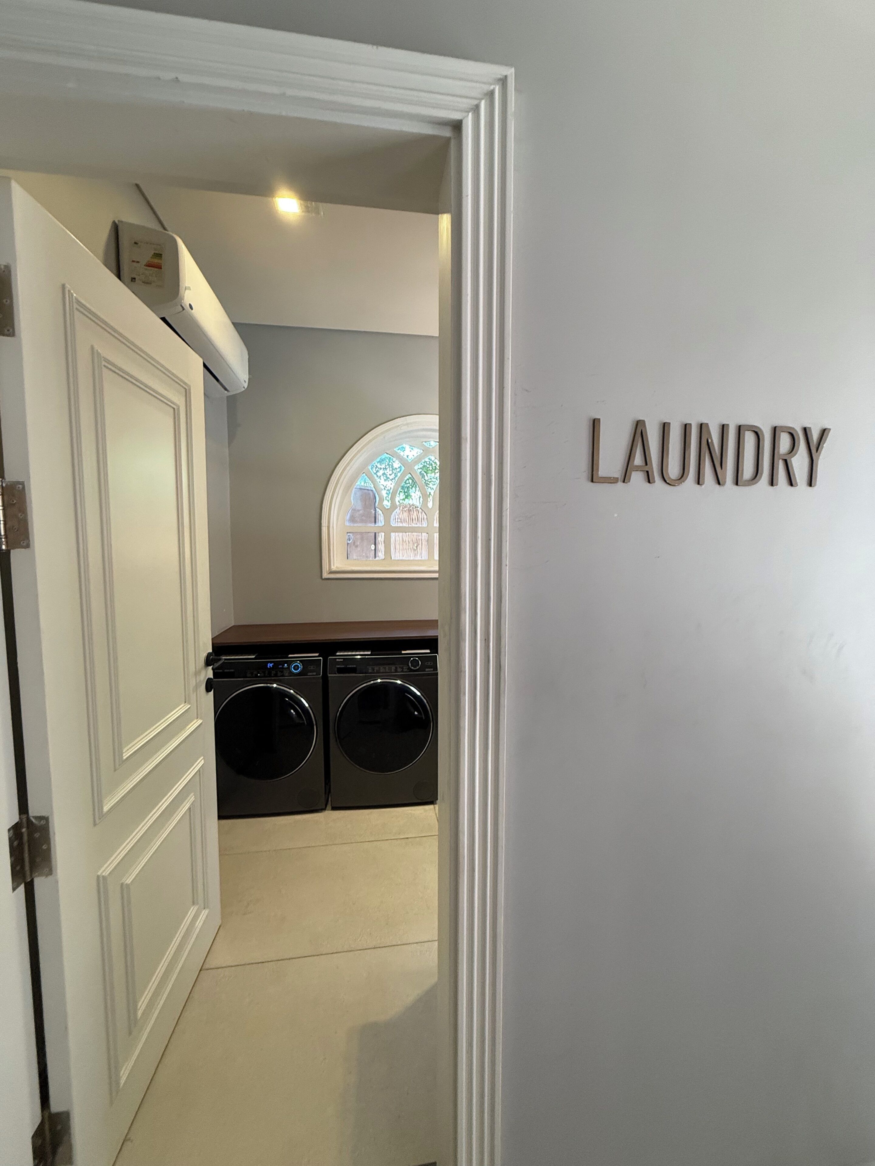 Uma lavanderia com duas máquinas de lavar, uma janela com design decorativo e uma porta que dá para outro cômodo.