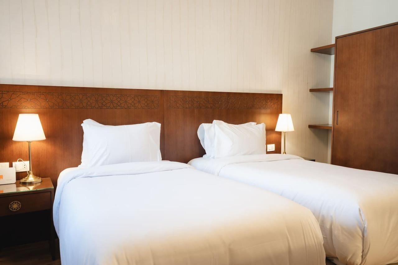 Quarto de hotel com duas camas, cabeceira de madeira, abajures e um guarda-roupa.