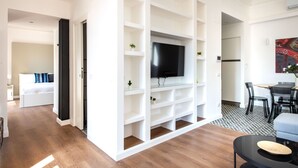 Uma sala de estar moderna com televisão em uma estante branca, uma área de jantar com mesa e cadeiras, e um quarto com cama e mesa de cabeceira.