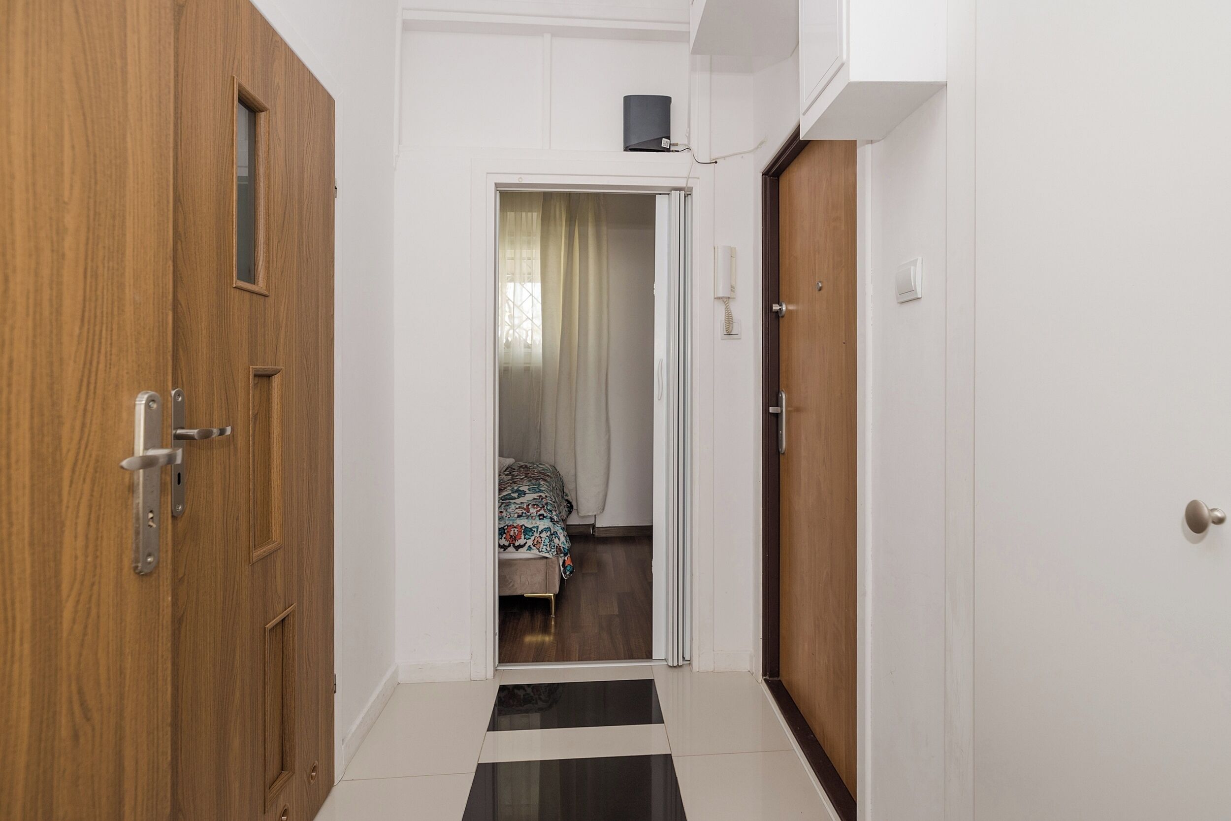 Um corredor estreito com uma porta de madeira à esquerda, uma porta branca à direita e uma cama visível por uma porta aberta ao fundo.