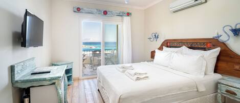 Quarto com cama, televisão, escrivaninha de madeira e vista para o mar através de uma porta de vidro.
