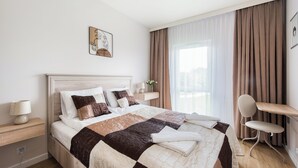 Uma cama bem arrumada com colcha, uma mesa de cabeceira com abajur, uma cadeira e uma janela com cortinas.