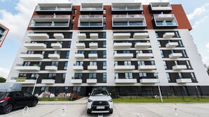 Um edifício residencial moderno de vários andares, com varandas, área de estacionamento e um número de contacto visível.