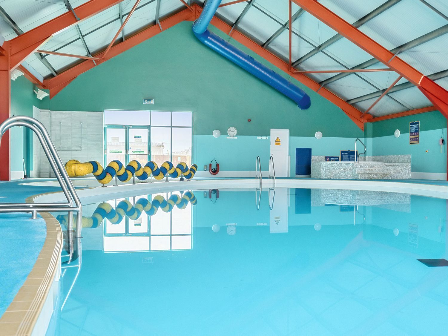 Piscina interna com teto alto, paredes de vidro transparente e painel de bolas azul e amarelo.