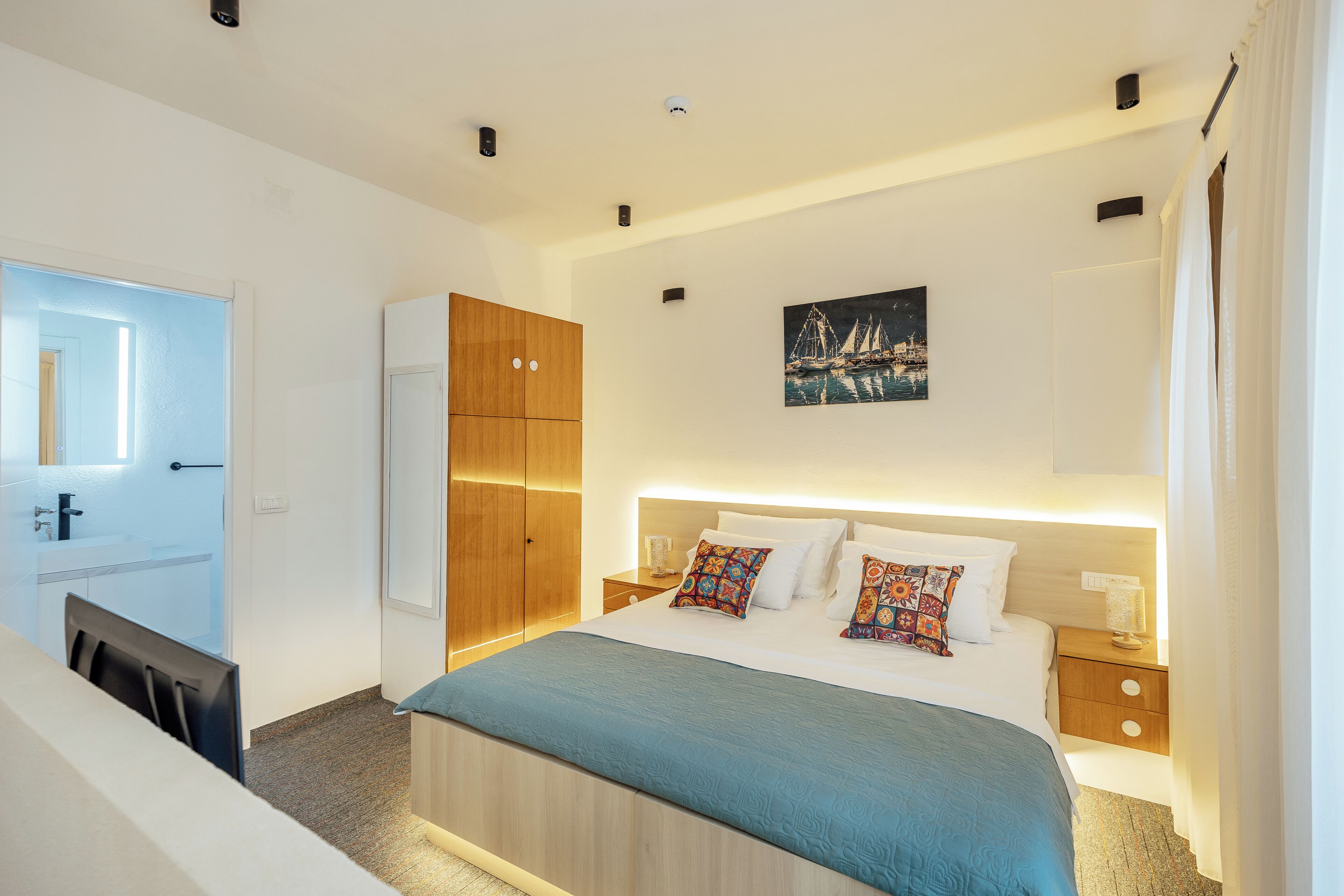 Quarto de hotel moderno com uma cama grande, um guarda-roupa de madeira, uma mesa de cabeceira com abajur e um quadro na parede.