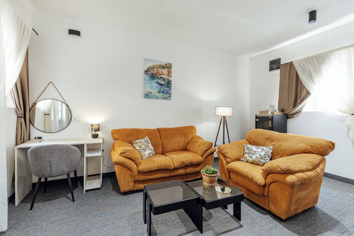 Um quarto com uma escrivaninha, dois sofás, uma poltrona, uma mesa de centro, um abajur, um quadro e uma janela com cortinas.