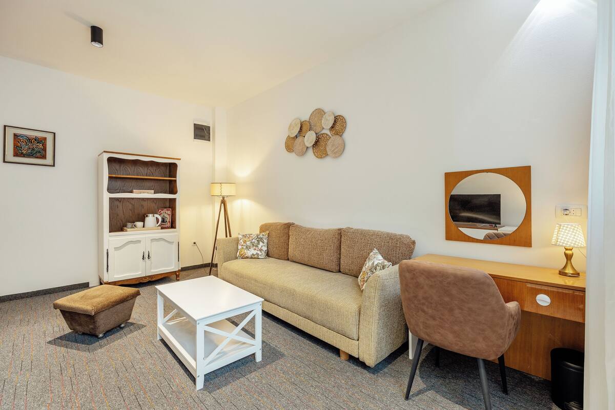 Quarto mobiliado com sofá, escrivaninha, cadeira, mesinha, armário, luminária, espelho e um enfeite de parede.