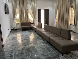 Uma sala de estar espaçosa com um sofá seccional bege, um pequeno sofá e uma mesa de centro. O cômodo tem piso de mármore e grandes janelas com cortinas bege.