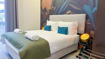 Cama bem arrumada com lençóis brancos e manta verde, mesa de cabeceira com abajur, e painel com folhagem tropical.