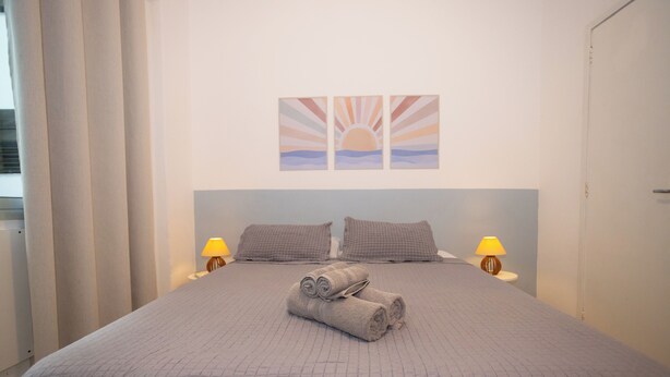 Cama bem arrumada com cobre acolchoado, duas almofadas e toalhas dobradas. Duas luminárias de cabeceira de cada lado da cama.