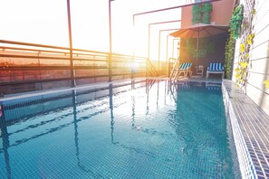 Uma piscina com água azul cristalina, um terraço ensolarado com grade e algumas cadeiras de descanso.