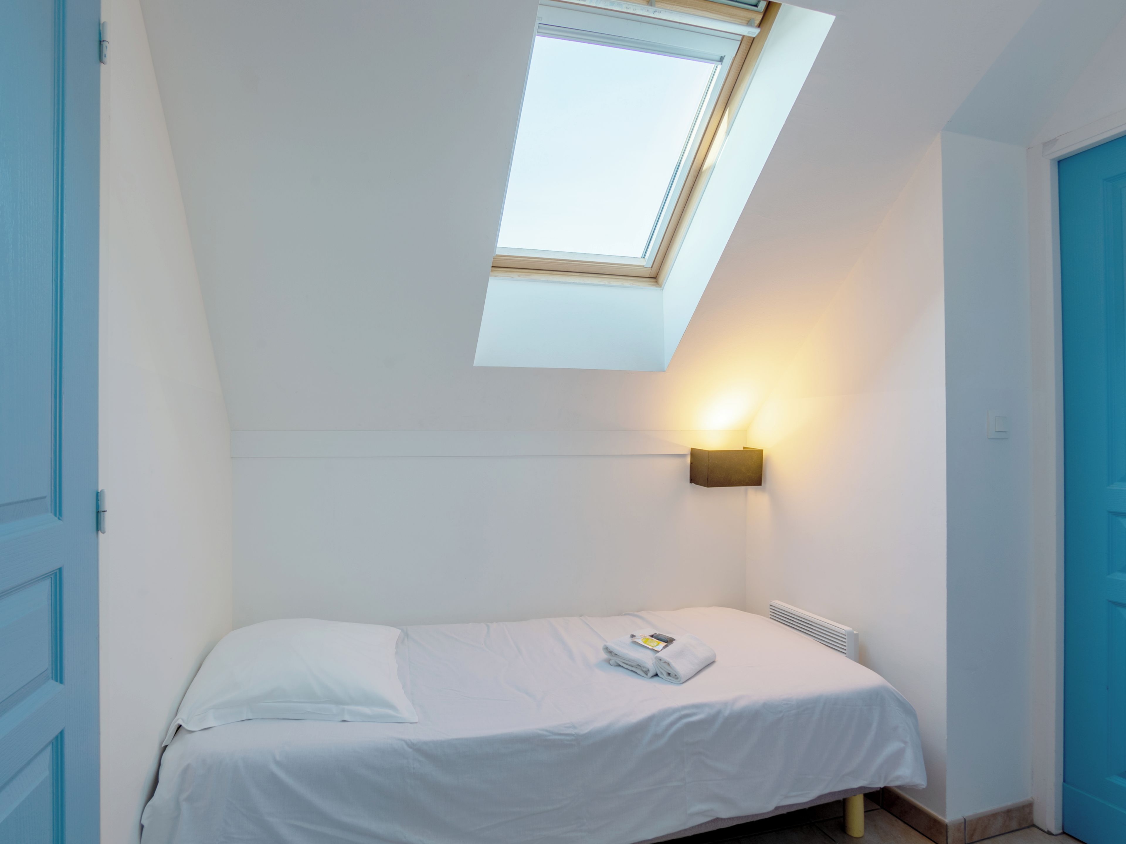 Quarto pequeno com uma cama de solteiro, claraboia, luminária na parede e radiador.
