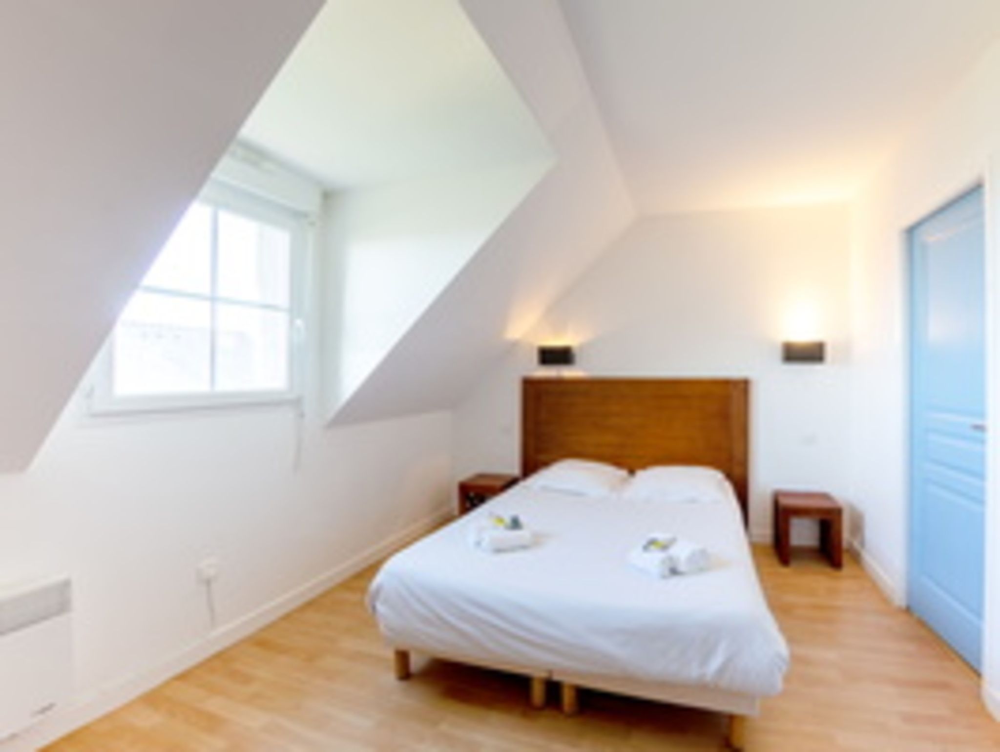 Quarto com teto inclinado, cama com almofadas e cobertor, mesas de cabeceira e uma janela.