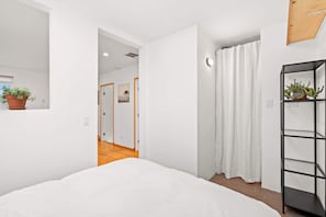 Quarto com cama, um vaso de planta, prateleira e porta que dá para outro cômodo.