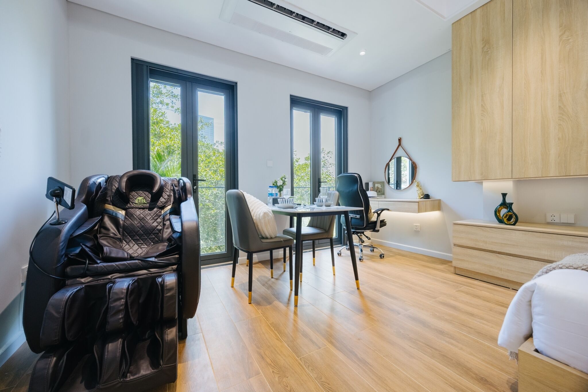Quarto moderno com cadeira de massagem, mesa de jantar com cadeiras e janelas amplas com vista para o verde.