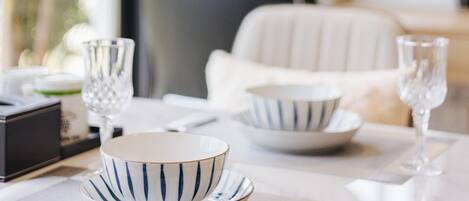 Uma mesa de jantar posta com tigelas brancas e listradas, uma colher e copos de vinho. Duas cadeiras, uma preta e outra branca, estão posicionadas atrás da mesa.