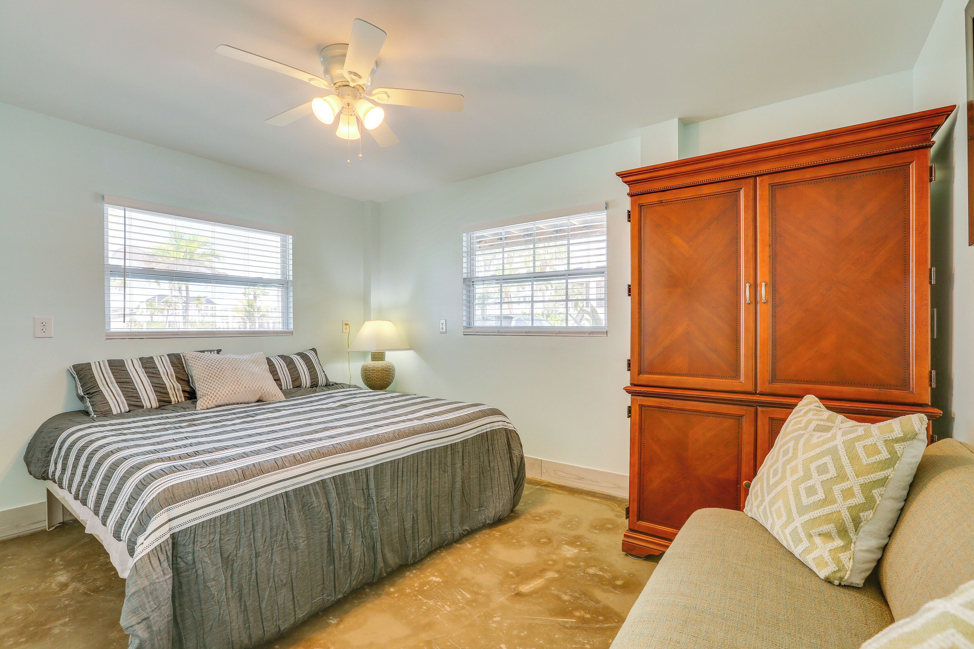 Quarto com cama, guarda-roupa de madeira, duas janelas e um ventilador de teto.