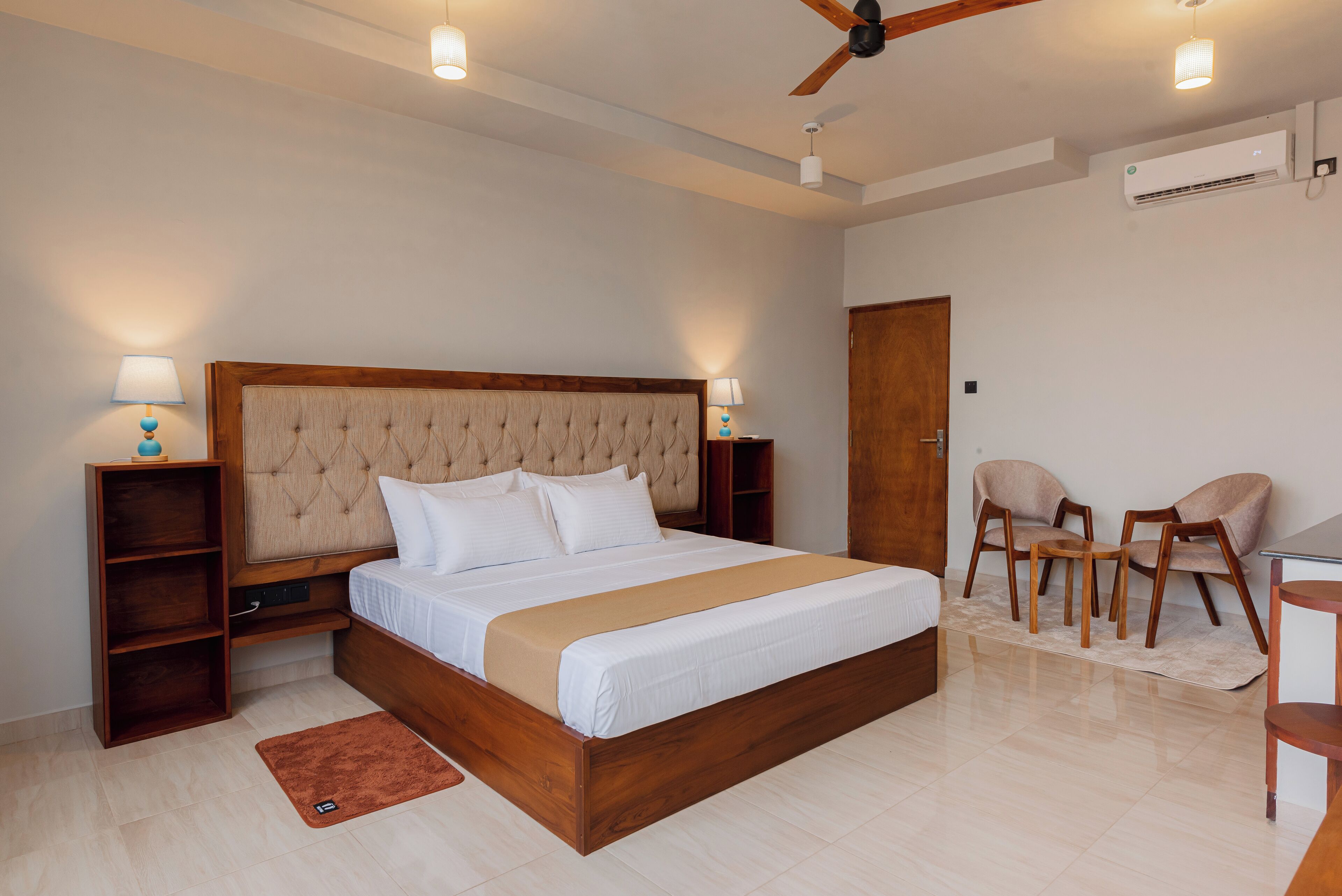 Quarto com uma cama grande, cabeceira de madeira, mesas de cabeceira com luminárias, ventilador de teto e um pequeno espaço de estar com cadeiras.