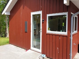 Um edifício vermelho com porta e janela emolduradas em branco, uma caixa de eletricidade visível e um pátio de concreto com um par de sapatos.